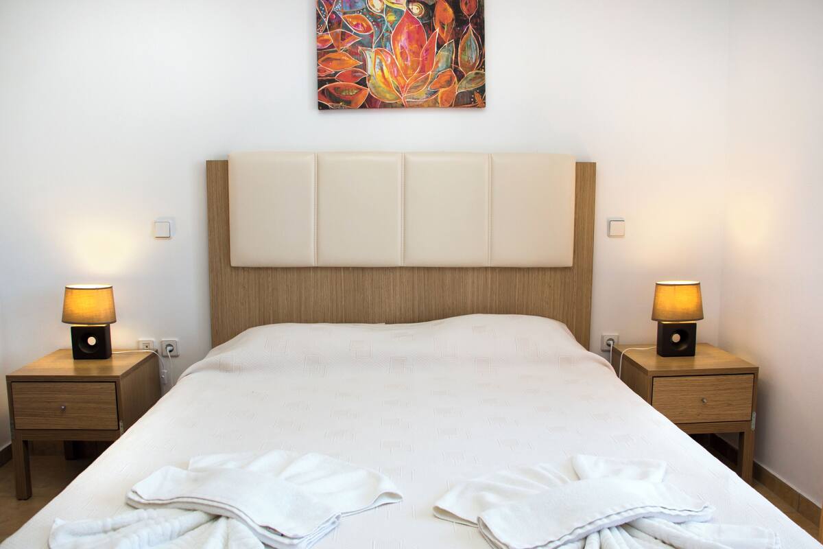 Cama bem arrumada com cabeceira, duas mesas de cabeceira com abajures e um quadro colorido na parede.