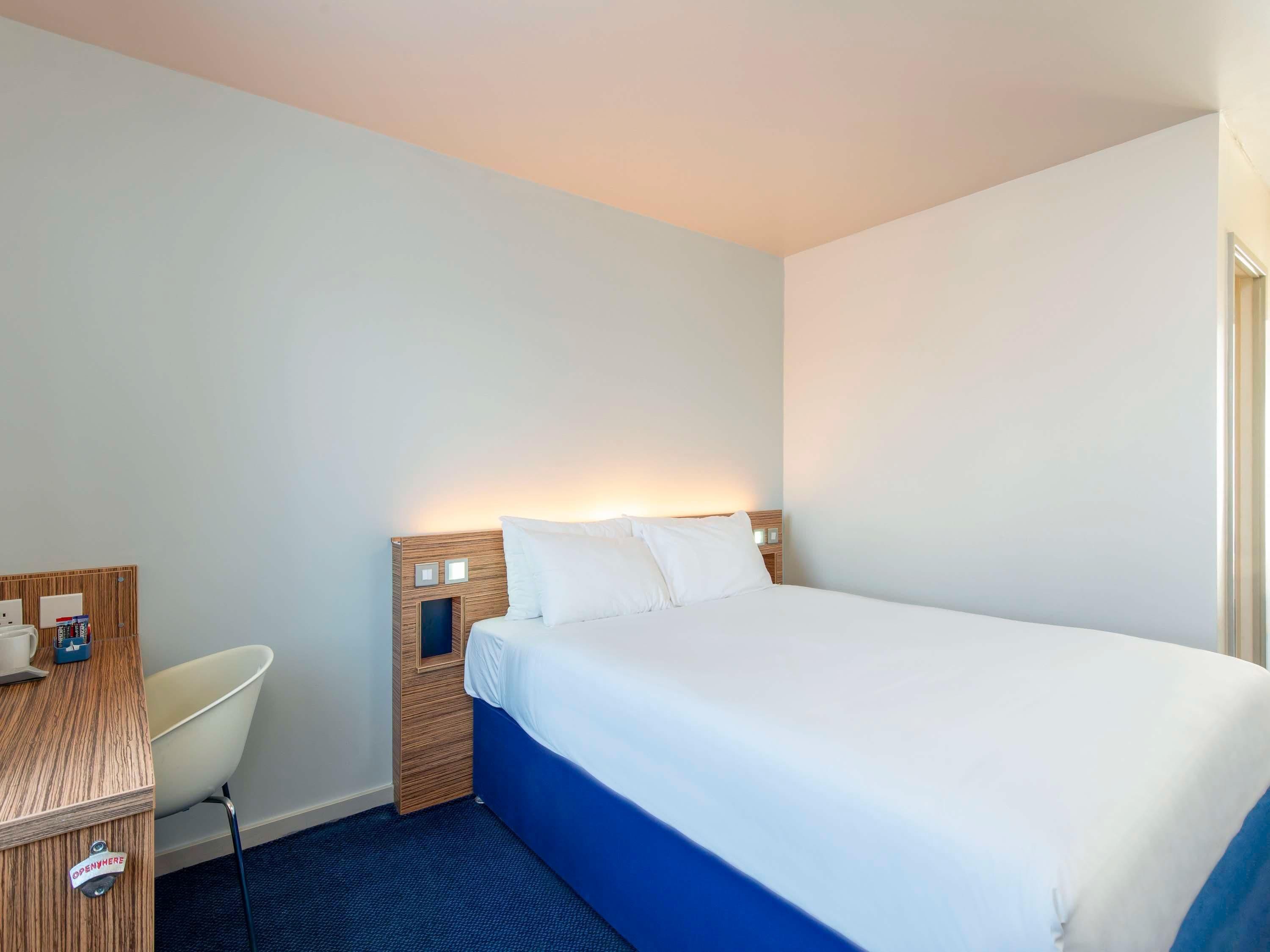 Quarto de hotel com cama, escrivaninha, cadeira e mesa de cabeceira.