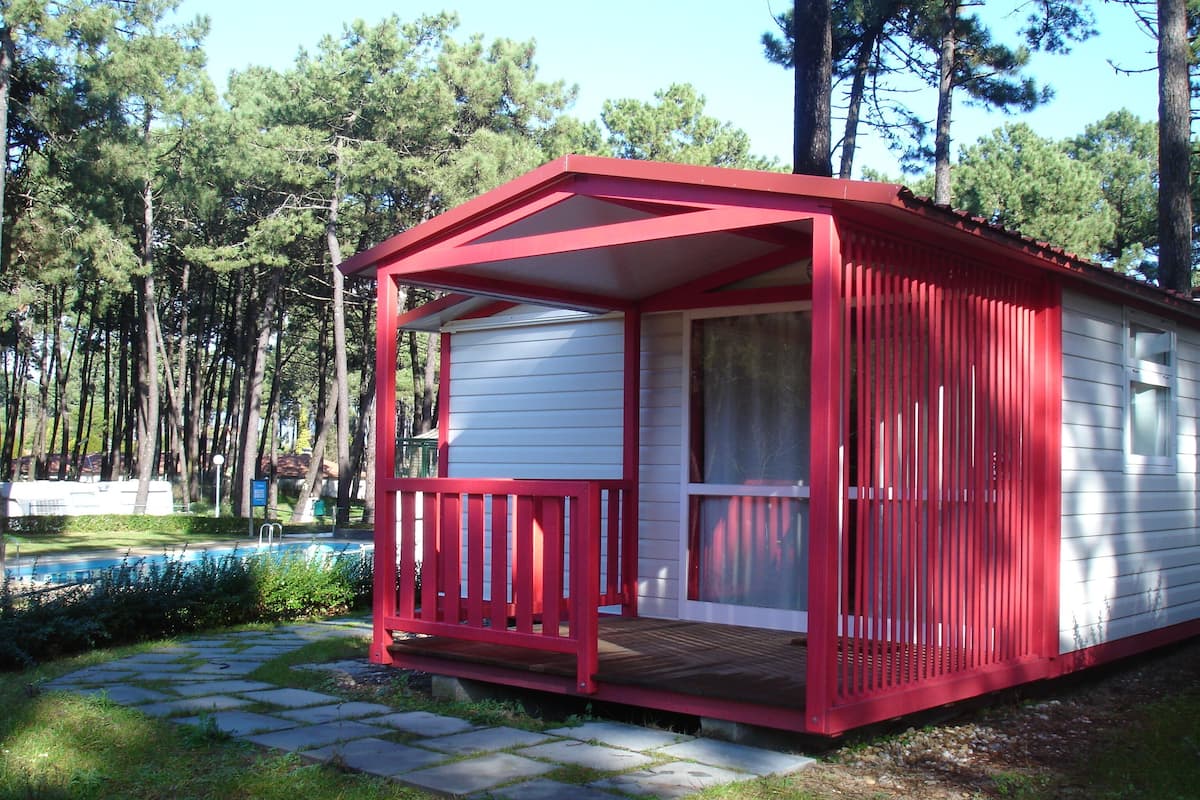 Uma cabana pequena, térrea, com fachada vermelha e varanda. Está rodeada por um caminho de pedra e vegetação exuberante.