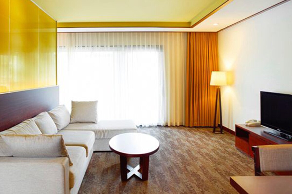 Quarto de hotel com sofá, mesa de centro redonda, televisão e luminária.
