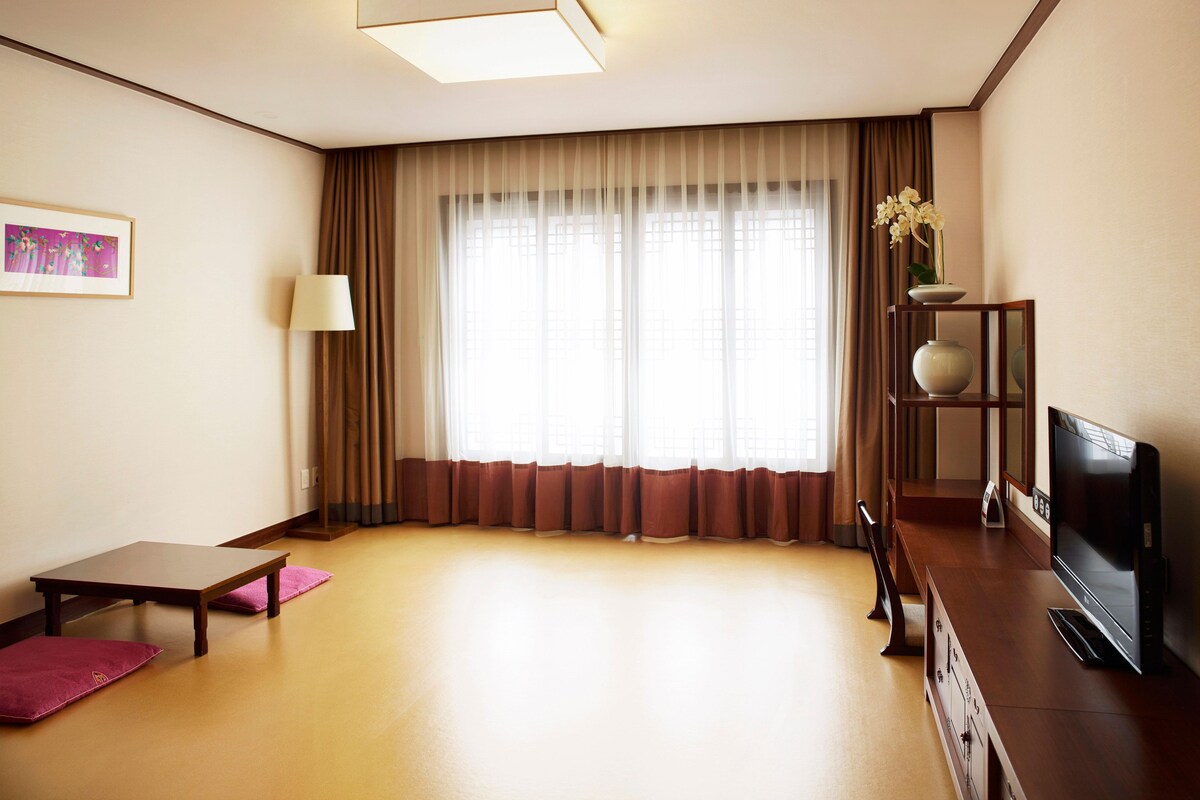 Quarto de hotel com uma grande janela, televisão de tela plana, escrivaninha de madeira e um pequeno aparador com um vaso.