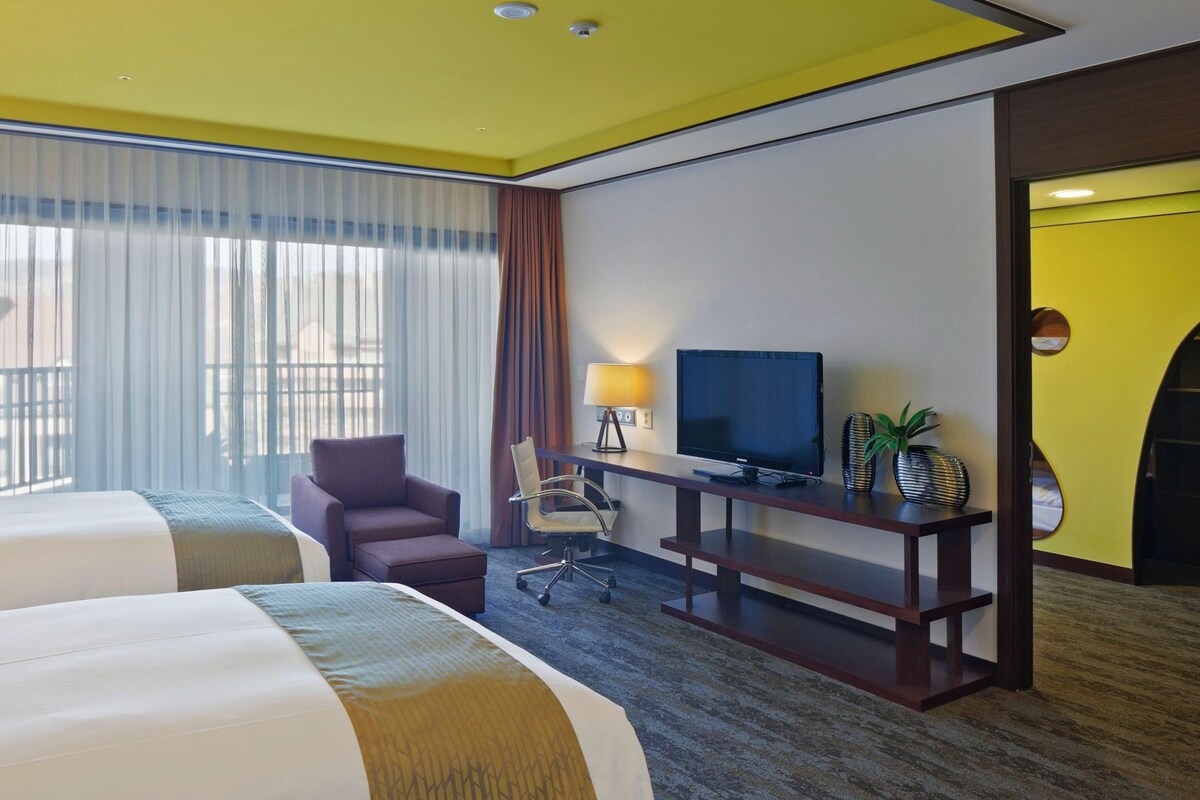 Quarto de hotel com duas camas, uma mesa de trabalho com televisão, uma cadeira e uma luminária de mesa.