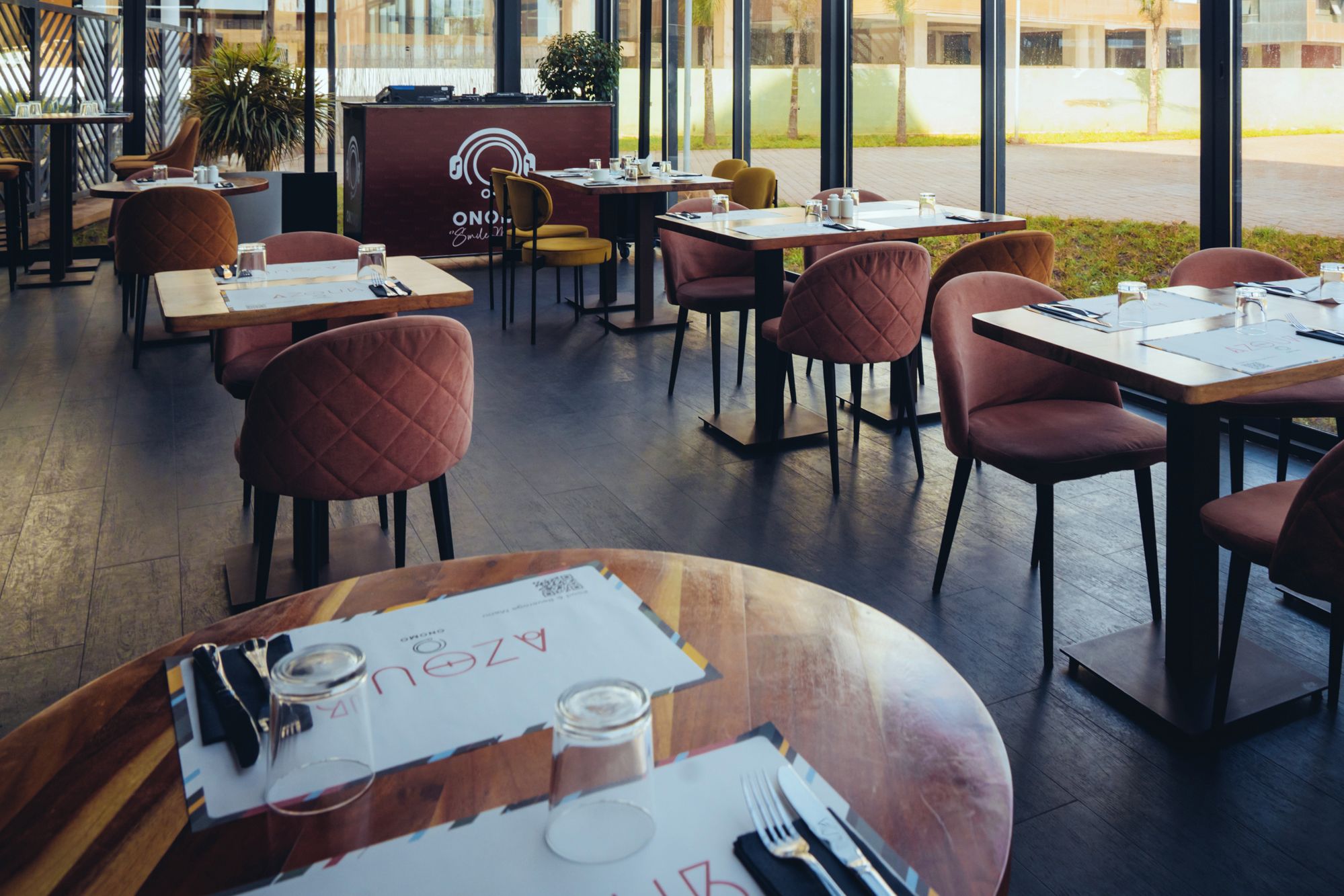 Restaurante com mesas e cadeiras de madeira, área de bar e grandes janelas com vista para a rua.