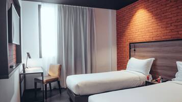 Quarto de hotel com duas camas, uma escrivaninha, uma cadeira e uma janela grande com cortinas.