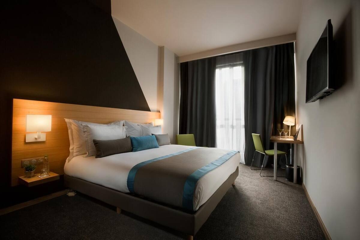 Quarto de hotel com cama, mesas de cabeceira, escrivaninha, cadeira, televisão e janela com cortinas.
