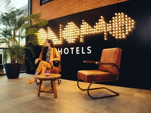 Uma pessoa sentada em uma cadeira de madeira com almofada, em frente a uma parede com a placa 'HOTELS' e uma luminária decorativa.