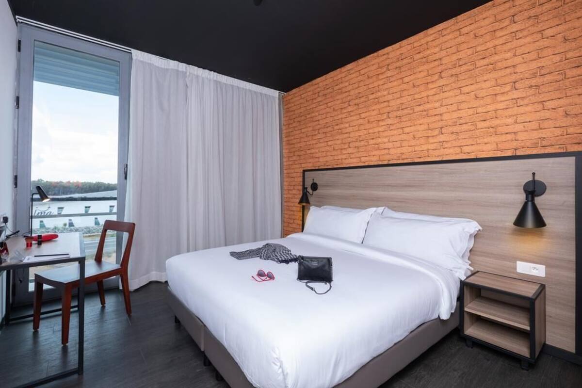 Quarto de hotel com uma cama grande, uma escrivaninha, uma cadeira e vista para a água através de uma janela.