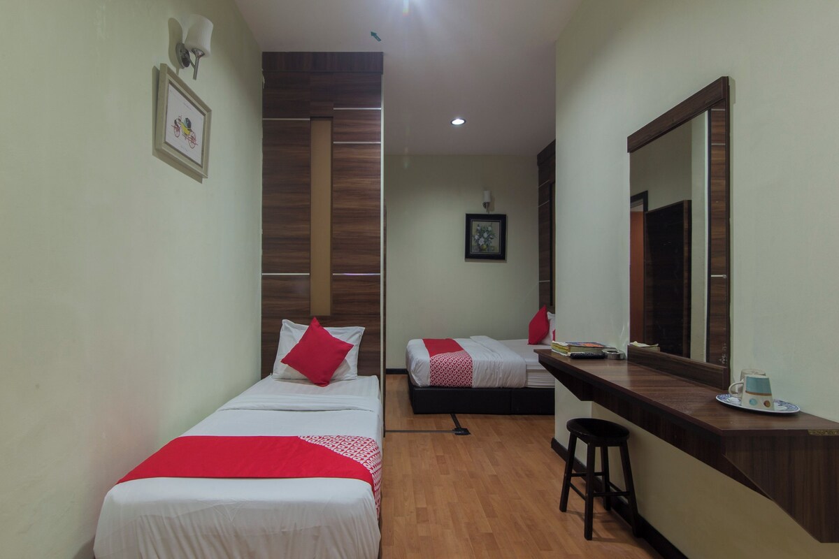Quarto de hotel com duas camas de solteiro, piso de madeira, uma pequena escrivaninha com espelho e uma área de cozinha compacta.