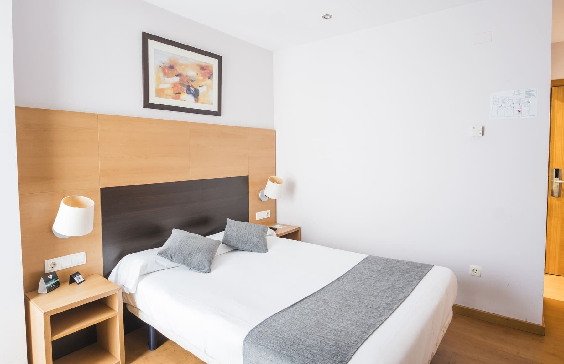 Quarto de hotel com uma cama, mesas de cabeceira, uma luminária na parede, um quadro emoldurado e uma porta.