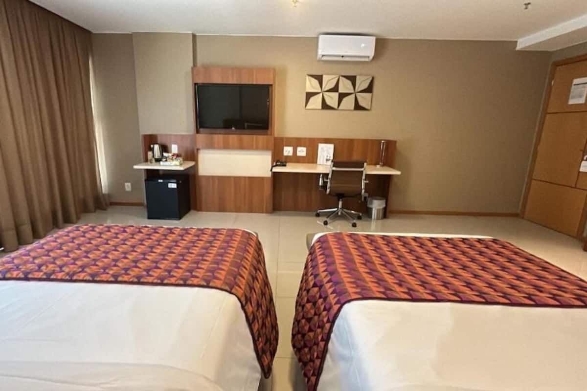 Quarto de hotel com duas camas, uma escrivaninha, uma cadeira, uma televisão e um guarda-roupa.