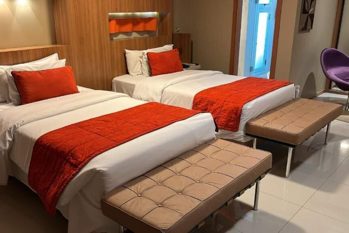 Quarto de hotel com duas camas, um banco, uma cadeira e uma mesinha de cabeceira.