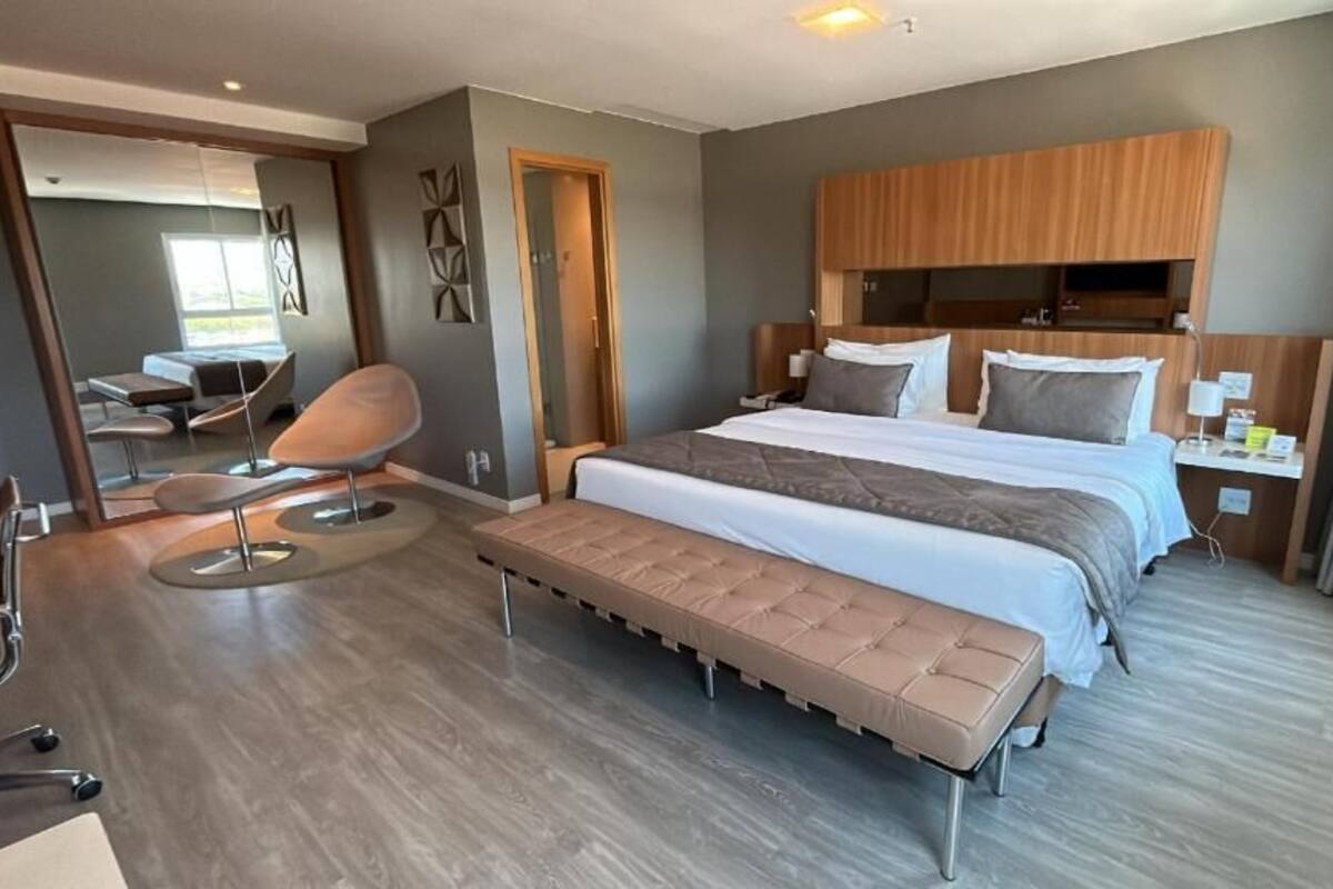 Quarto de hotel moderno com uma cama grande, área de estar com uma poltrona, escrivaninha e uma pequena mesa.