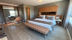 Quarto de hotel moderno com uma cama grande, área de estar com uma poltrona, escrivaninha e uma pequena mesa.