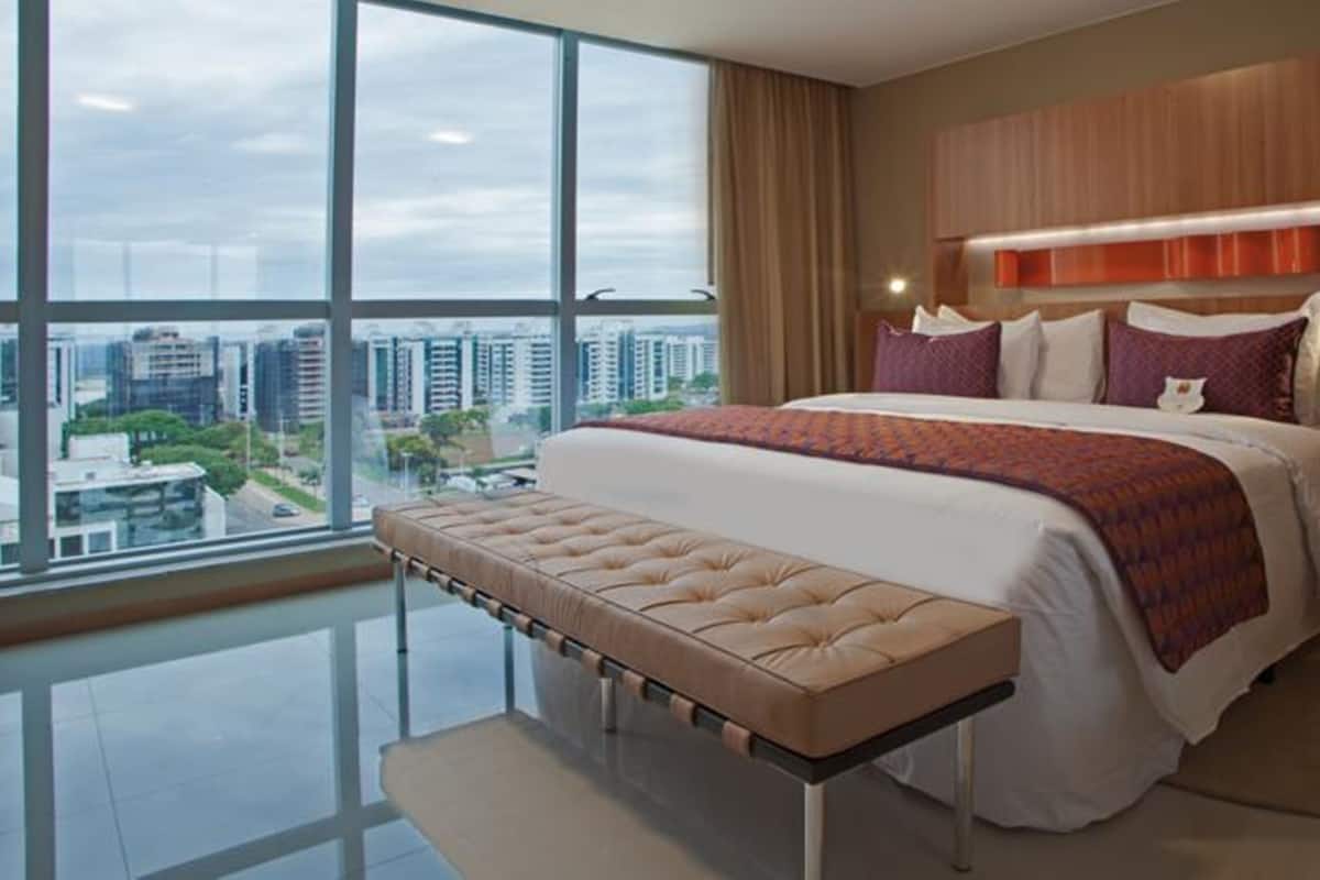 Quarto de hotel moderno com uma cama grande, um banco, uma mesa de cabeceira e vista para a cidade.