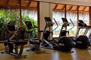 Ginásio com bicicletas ergométricas, elípticos e esteiras, com grandes janelas dando vista para uma piscina e vegetação exuberante.