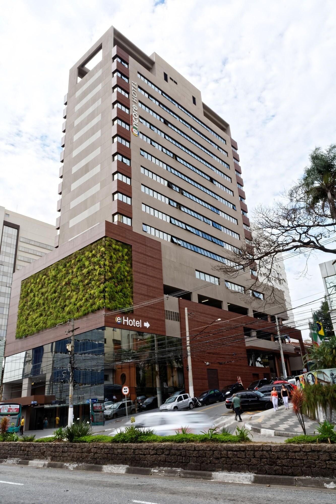 Hotel de vários andares com design moderno, apresentando um jardim vertical verdejante em uma lateral e uma placa visível com o nome do hotel.
