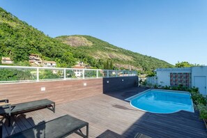 Uma área de piscina no terraço com um deck de madeira, piscina e vista para uma colina com edifícios.