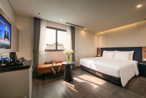 Quarto de hotel com cama, sofá, uma mesinha com um vaso de flores, espaço para café e uma televisão exibindo notícias.