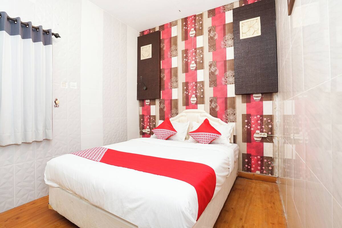 Cama bem arrumada com colcha vermelha e branca, piso de madeira e parede estampada com dois relógios pendurados.