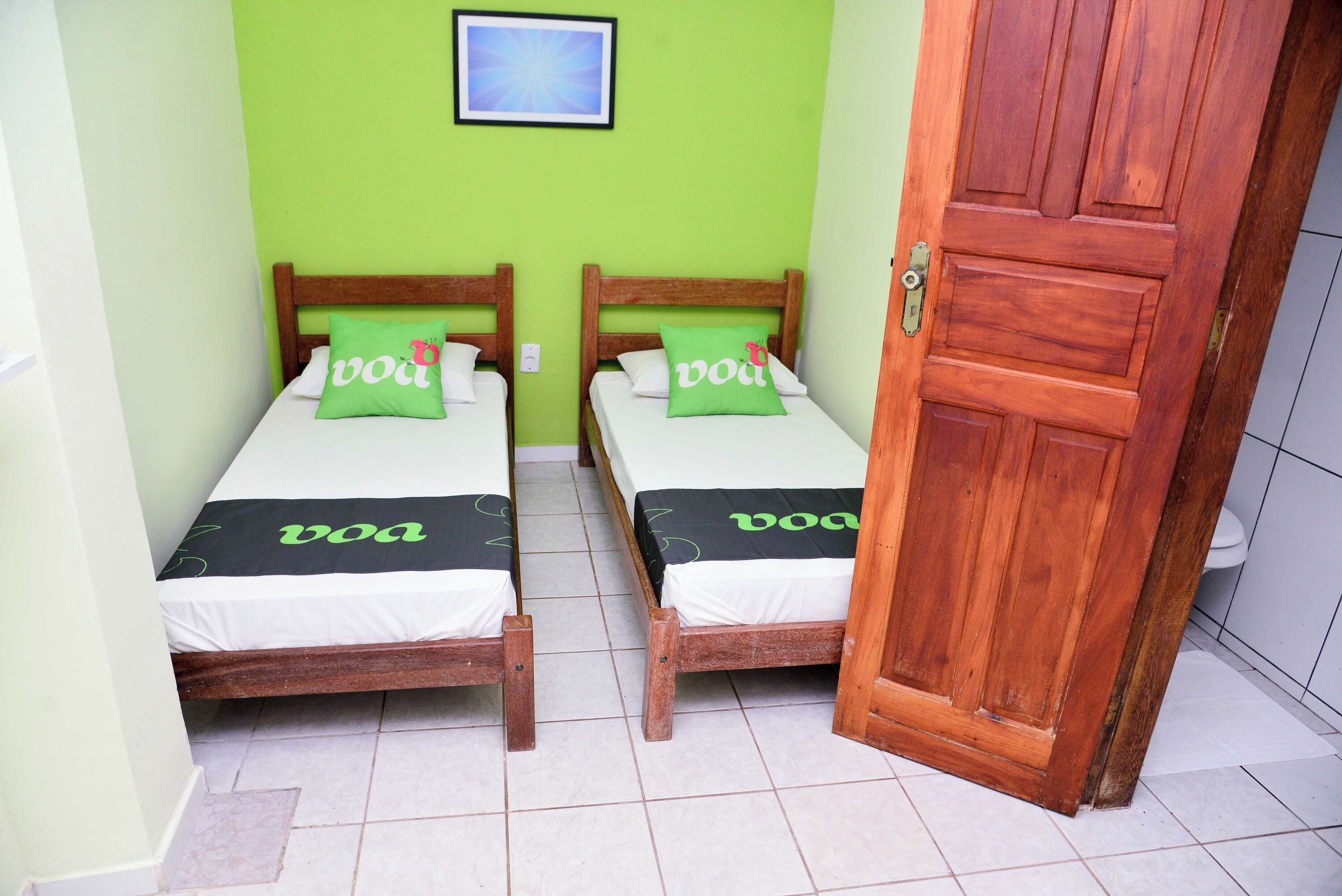Quarto com duas camas de madeira, cada uma com travesseiro verde e colchão branco, e uma porta de madeira.