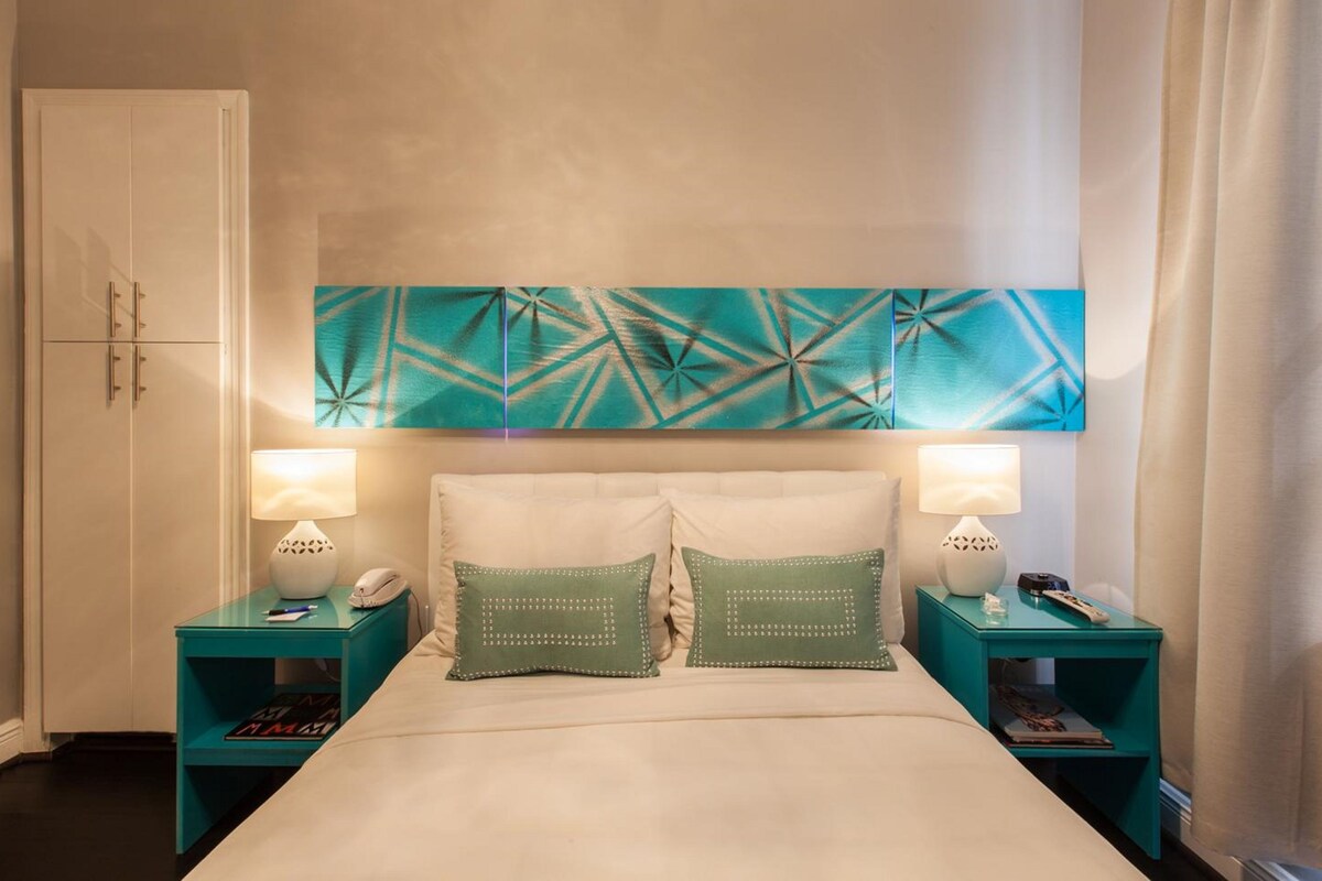Quarto de hotel com cama, duas mesas de cabeceira, um guarda-roupa branco e uma obra de arte geométrica na parede.