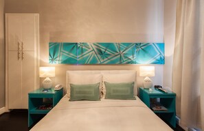 Quarto de hotel com cama, duas mesas de cabeceira, um guarda-roupa branco e uma obra de arte geométrica na parede.