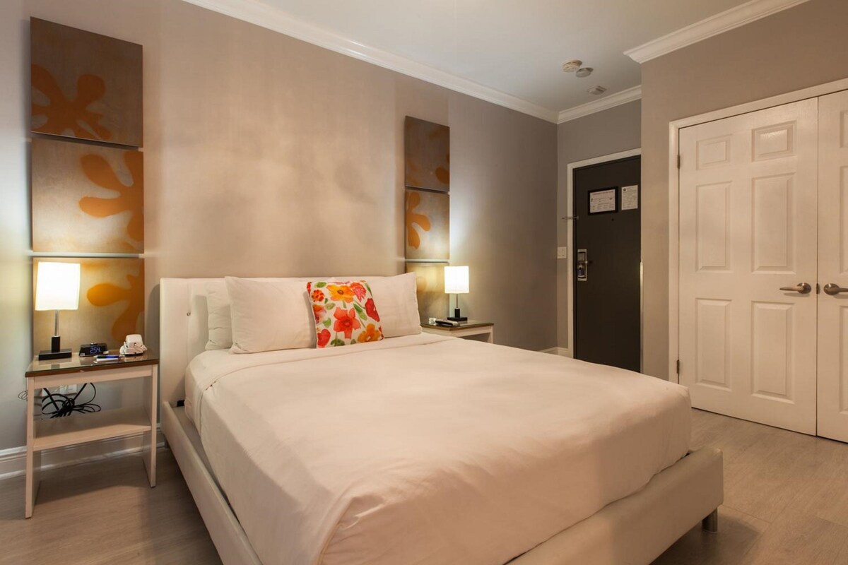 Quarto de hotel com cama, mesas de cabeceira, luminária, porta e parede decorada com arte abstrata.