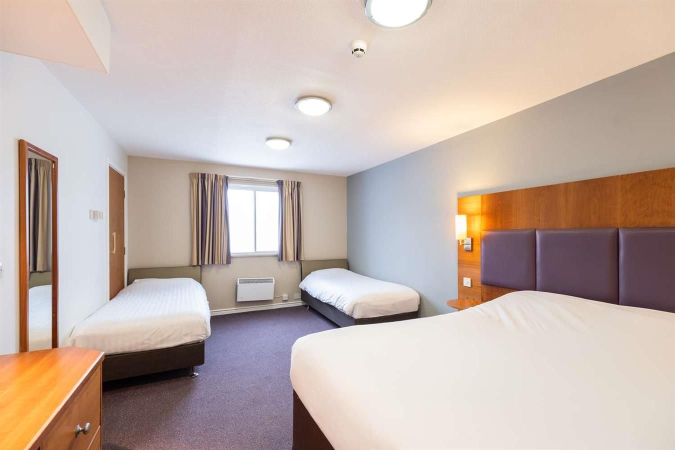 Quarto de hotel com duas camas de solteiro, penteadeira de madeira, janela com cortinas, luminária de teto e abajur na parede.