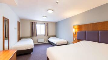 Quarto de hotel com duas camas de solteiro, penteadeira de madeira, janela com cortinas, luminária de teto e abajur na parede.