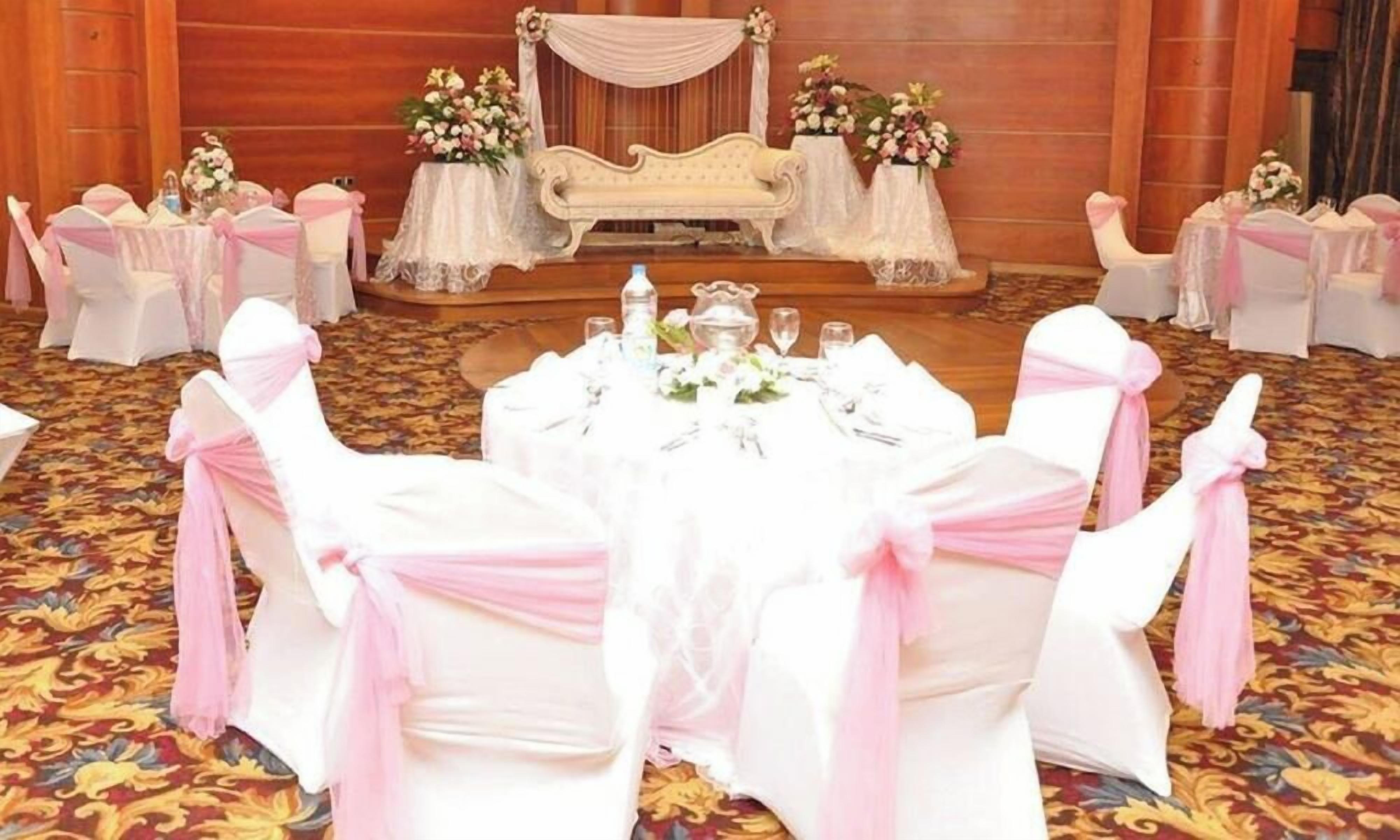 Salão de festas com mesas redondas dispostas para um evento formal, com palco central, sofá e arranjos florais.