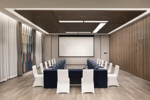 Uma sala de conferências com uma mesa comprida, cadeiras brancas e uma tela de projeção.