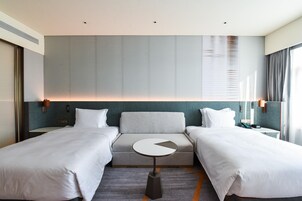 Quarto de hotel com duas camas, um sofá, uma mesa redonda pequena e uma janela grande com cortinas.