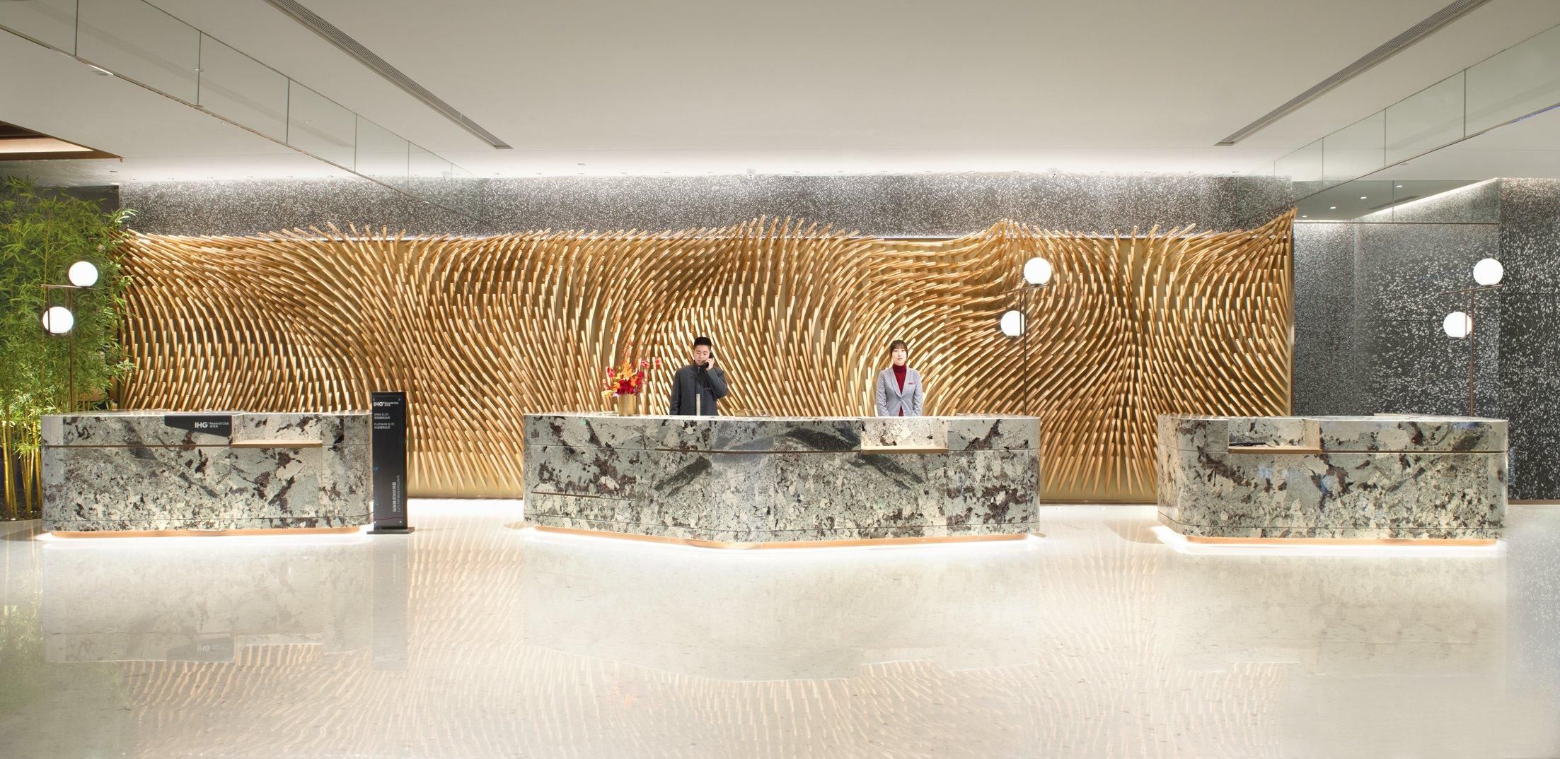 Uma área de recepção moderna com três balcões de mármore, parede com padrão de bambu e guarda-corpo de vidro.