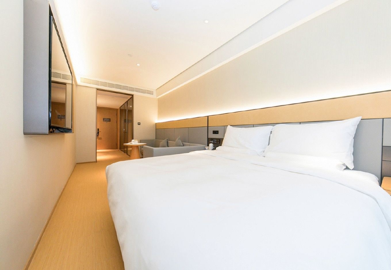 Quarto de hotel moderno com uma cama grande, mesa de cabeceira, TV de tela plana na parede e um espaço de estar separado com um sofá e poltronas.