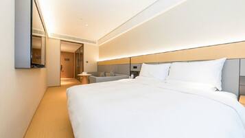Quarto de hotel moderno com uma cama grande, mesa de cabeceira, TV de tela plana na parede e um espaço de estar separado com um sofá e poltronas.