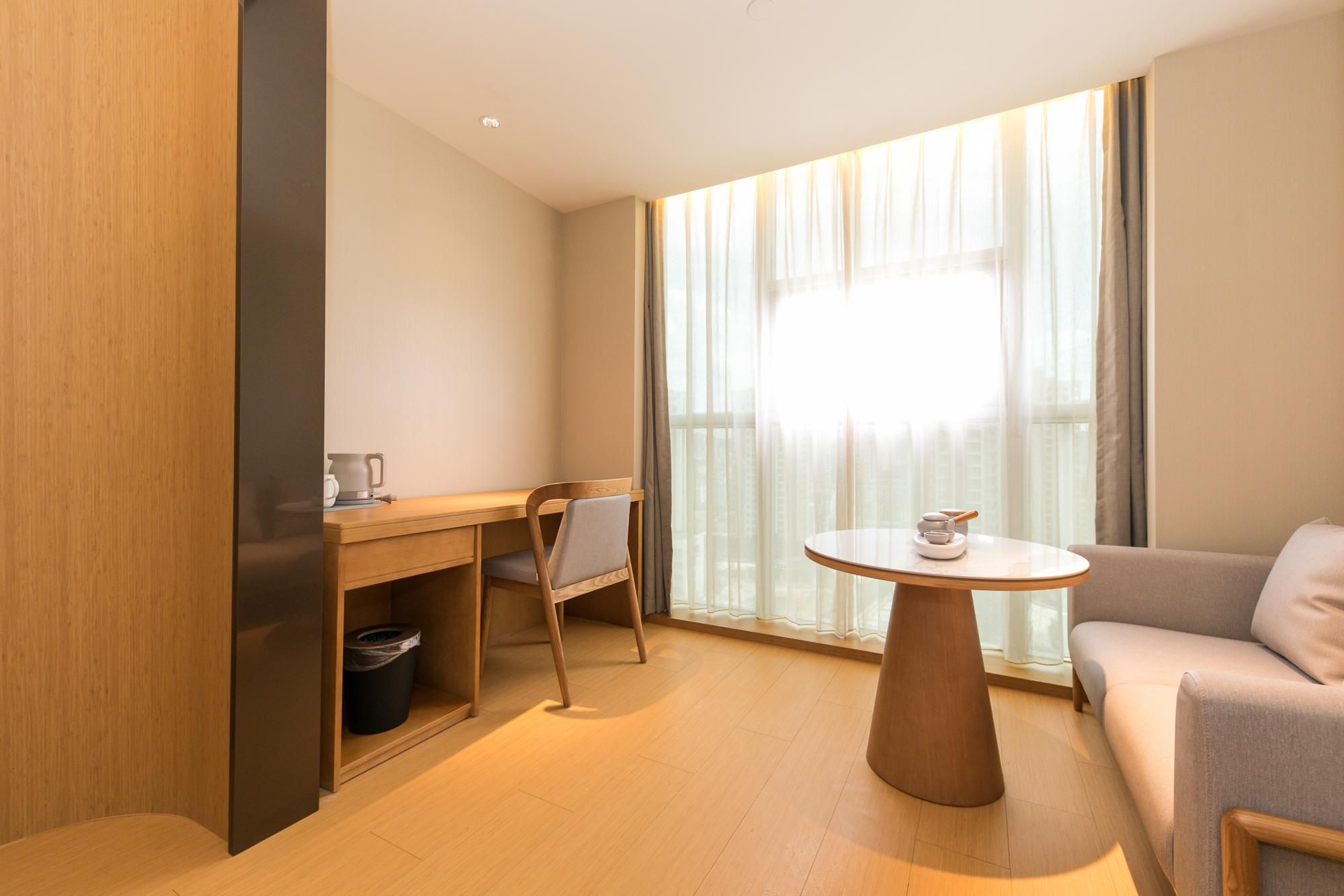 Quarto de hotel moderno com escrivaninha de madeira, uma cadeira, uma mesa redonda com uma xícara, um sofá e uma janela grande com cortinas leves.