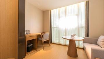 Quarto de hotel moderno com escrivaninha de madeira, uma cadeira, uma mesa redonda com uma xícara, um sofá e uma janela grande com cortinas leves.