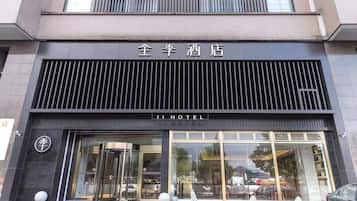 Entrada de um hotel com fachada moderna, grandes janelas de vidro e uma placa com o nome 'JI HOTEL'.