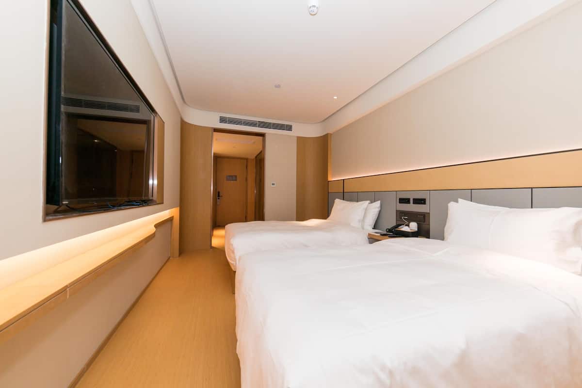 Quarto de hotel com duas camas, uma mesa de cabeceira, uma janela e uma porta que dá para outro espaço.