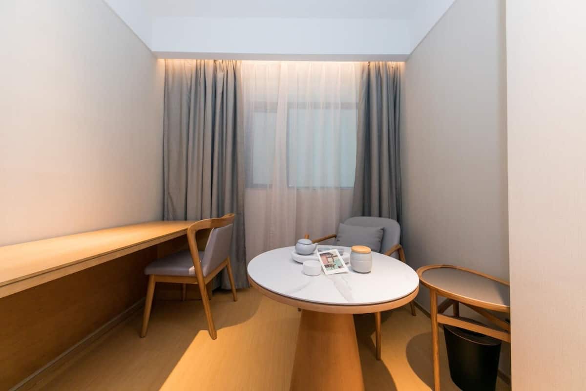 Quarto pequeno e moderno com uma mesa redonda, duas cadeiras, escrivaninha de madeira, lixeira e janela com cortinas.