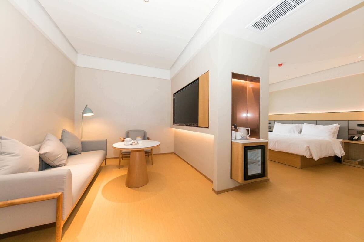 Quarto de hotel moderno com sofá, mesa de jantar, televisão, cama e um pequeno armário.