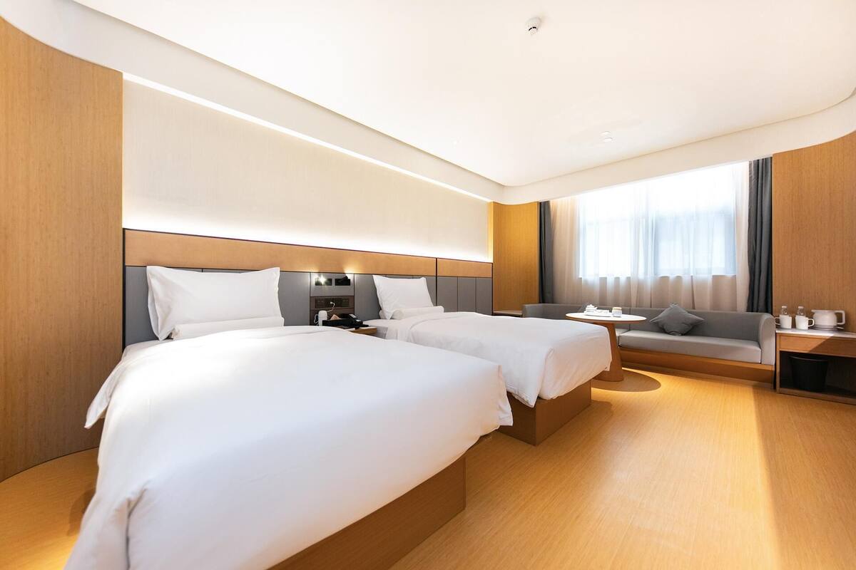 Quarto de hotel com duas camas, um sofá, uma escrivaninha e uma janela grande com cortinas.