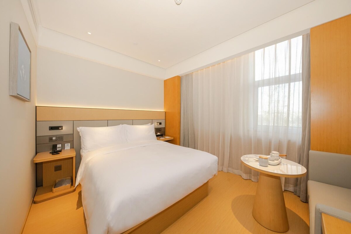 Quarto de hotel com uma cama grande, mesas de cabeceira, uma pequena mesa redonda e uma janela com cortinas.