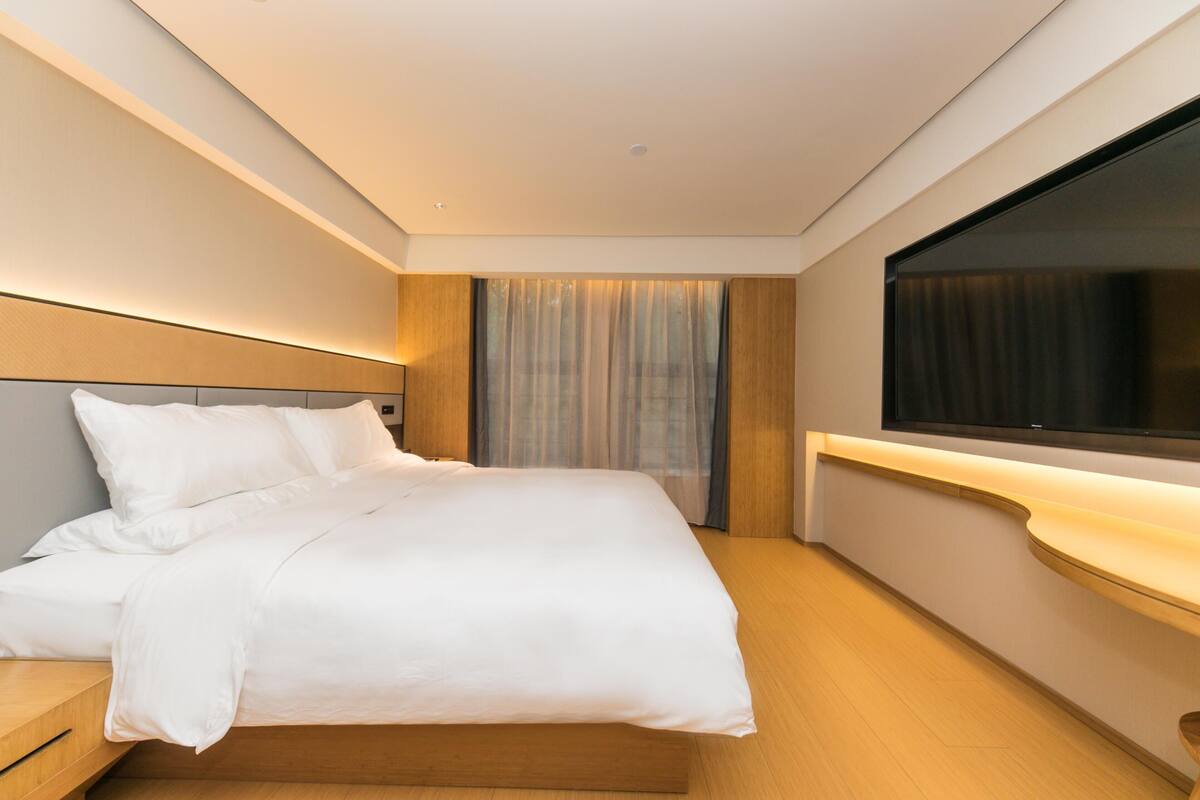 Quarto de hotel moderno com cama, mesa de cabeceira, uma janela grande com cortinas e uma TV de tela plana na parede.