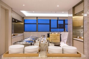 Buffet montado com várias estações de serviço, uma grande janela com vista e uma cozinha moderna ao fundo.