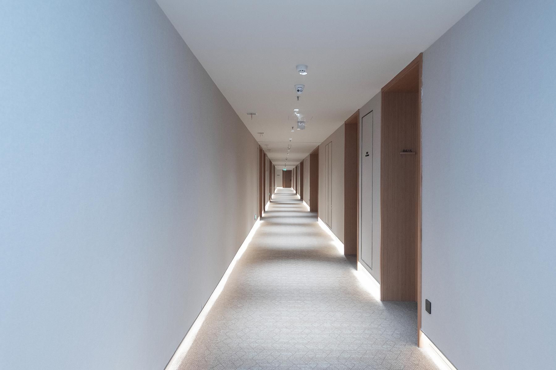 Um corredor comprido e estreito com piso coberto por carpete, iluminação embutida e portas em ambos os lados.