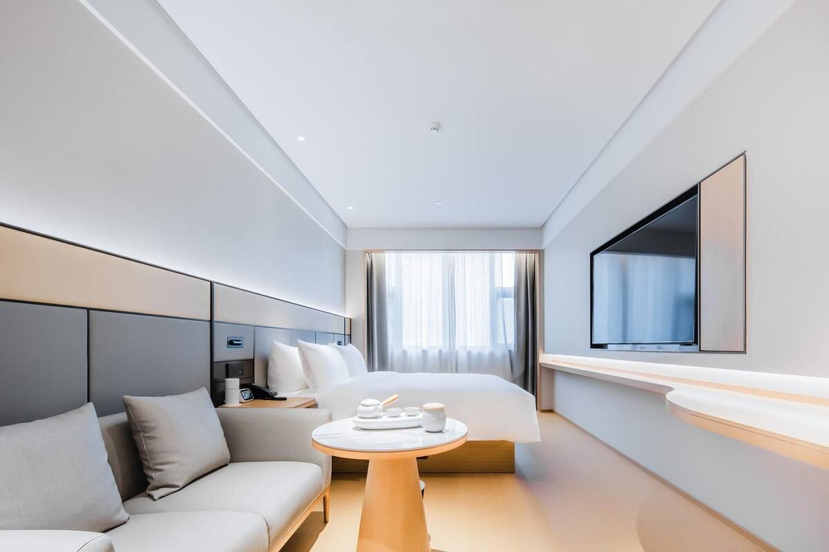 Quarto de hotel moderno com cama, sofá, mesinha com amenidades, televisão e uma grande janela com cortinas.