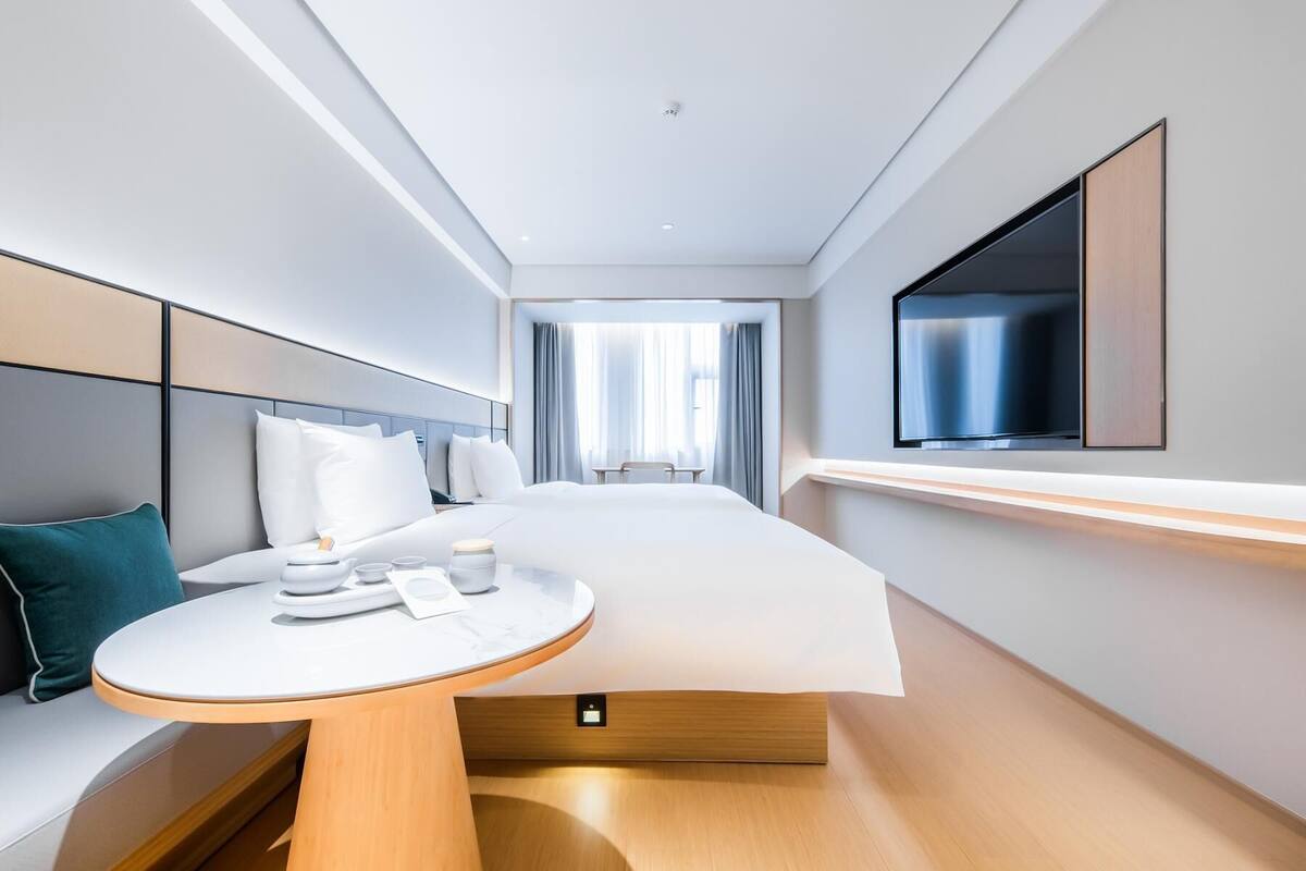 Quarto de hotel moderno com cama, mesa de cabeceira, televisão e vista para o corredor.