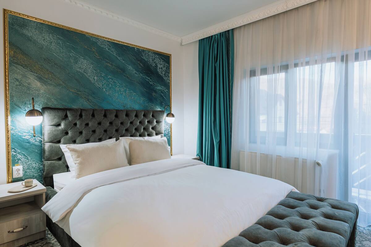 Quarto de hotel com uma cama grande, cabeceira estofada, mesa de cabeceira com abajur e uma grande pintura abstrata na parede.