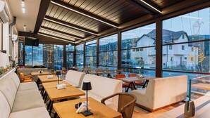 Um restaurante moderno com mesas de madeira e sofás brancos, grandes janelas com vista para edifícios e um corpo de água.